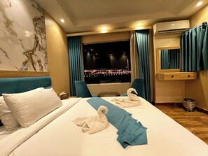 Quarto de hotel com cama, duas cadeiras, janela com cortinas, ar condicionado e prateleira na parede.