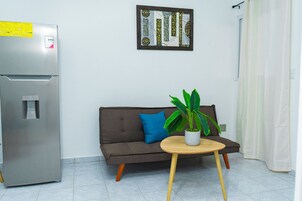 Uma sala de estar com sofá, uma mesinha redonda com planta em vaso, geladeira e uma obra de arte na parede.