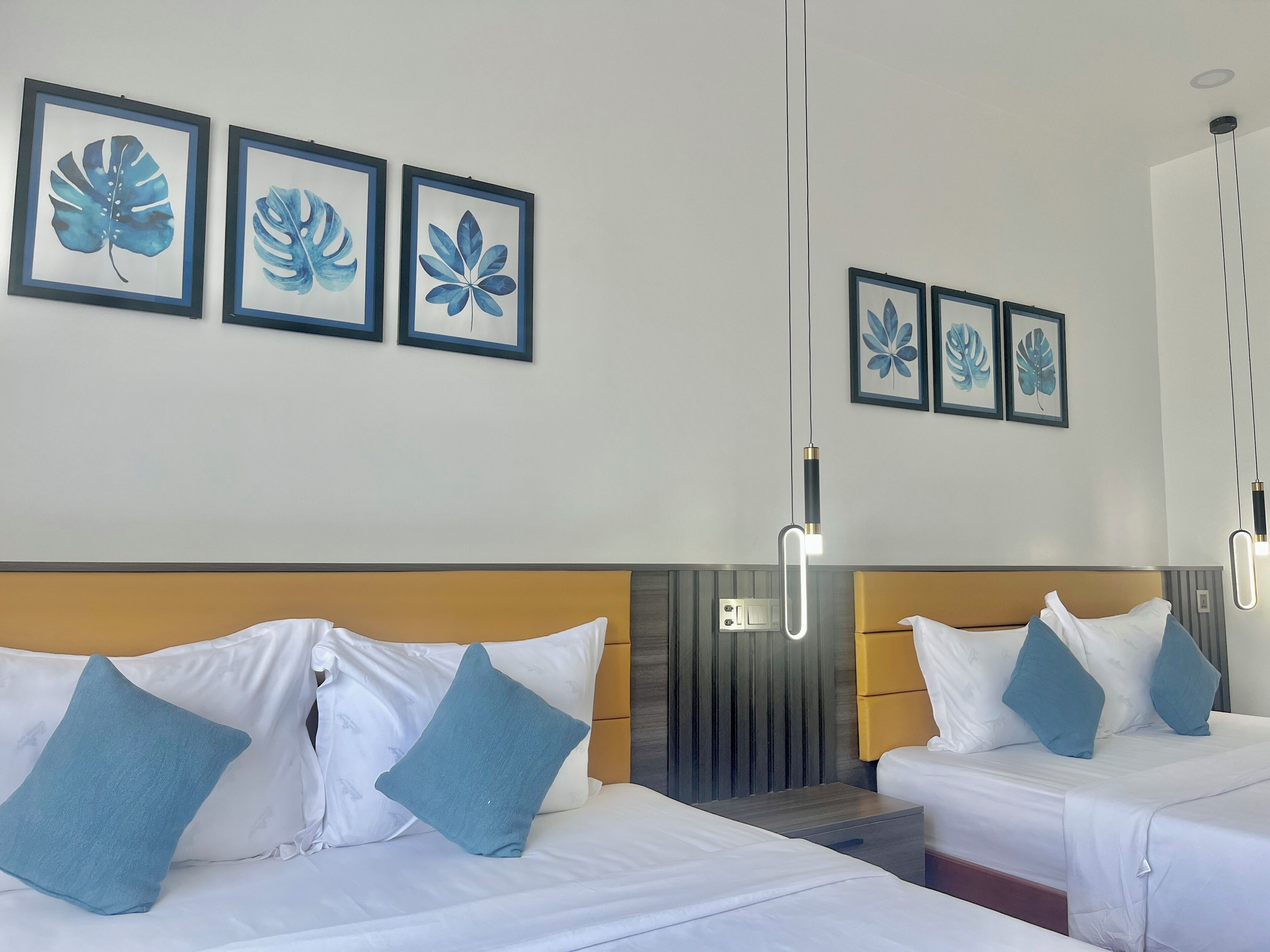 Quarto de hotel com duas camas, cada uma com roupa de cama branca e almofadas azuis. Há quadros com folhagens na parede.