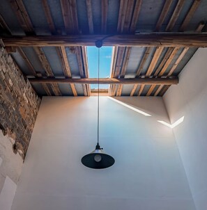 Quarto com teto inclinado, vigas de madeira aparentes, luminária pendente e vista do céu pelo teto.