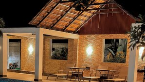 Uma piscina com guarda-corpo de vidro, cadeiras de descanso e um edifício com telhado telhado ao fundo.