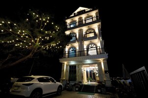 Um hotel de vários andares com uma entrada imponente, uma varanda no andar superior e um carro branco estacionado na frente.