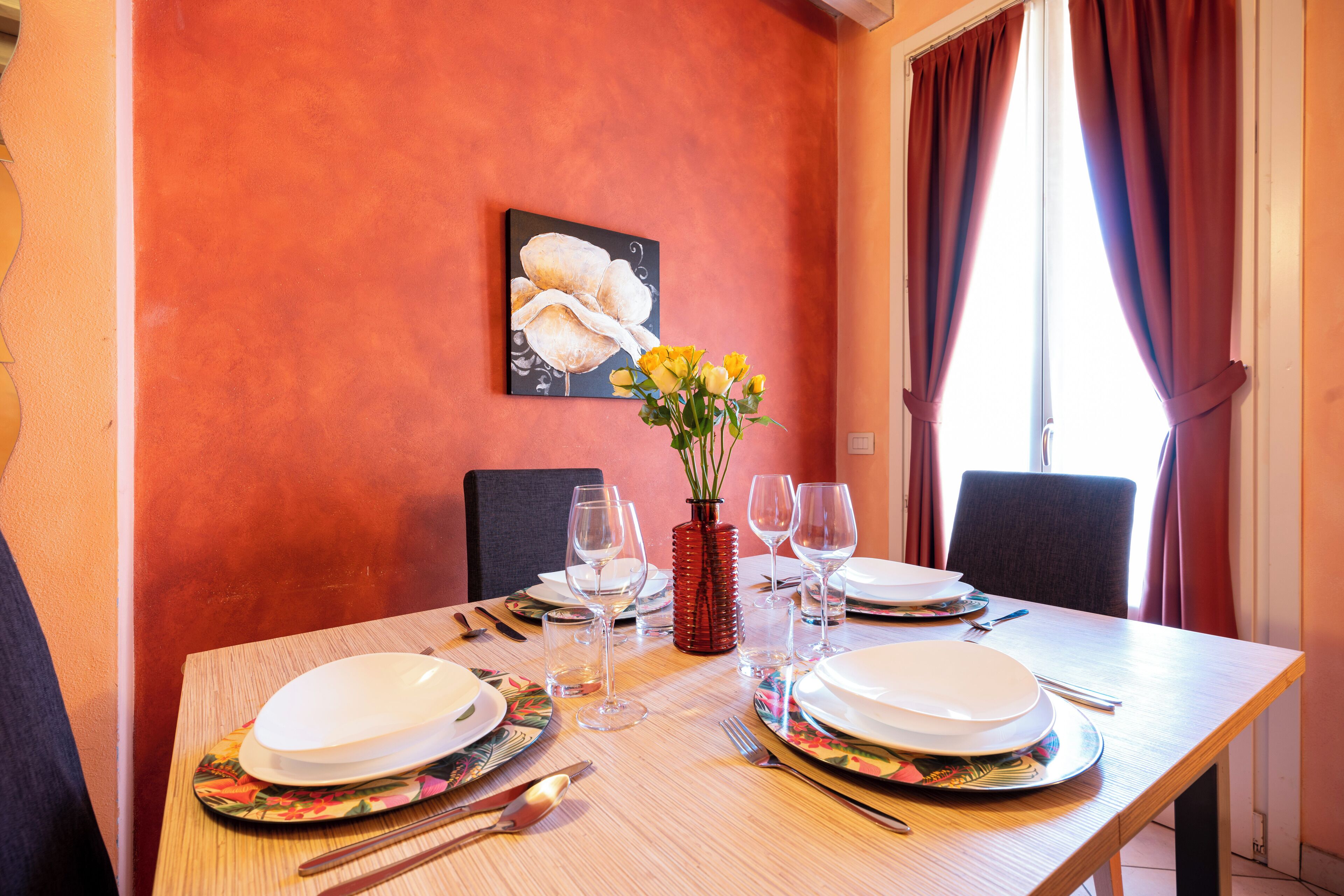 Uma mesa de jantar posta com pratos, copos e talheres, um vaso com flores e um quadro na parede.