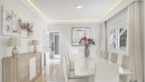 Uma sala de jantar moderna com uma mesa comprida, cadeiras brancas e um vaso de flores. Há um aparador de madeira com luminárias à esquerda e um quadro na parede.