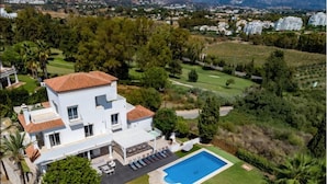 Uma casa grande e moderna com piscina, rodeada por vegetação exuberante e montanhas ao fundo.