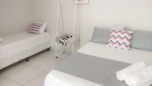 Quarto com duas camas, uma com travesseiro cinza e branco estampado e outra com travesseiro rosa e cinza em zigue-zague. Há uma toalha branca na cama em primeiro plano.