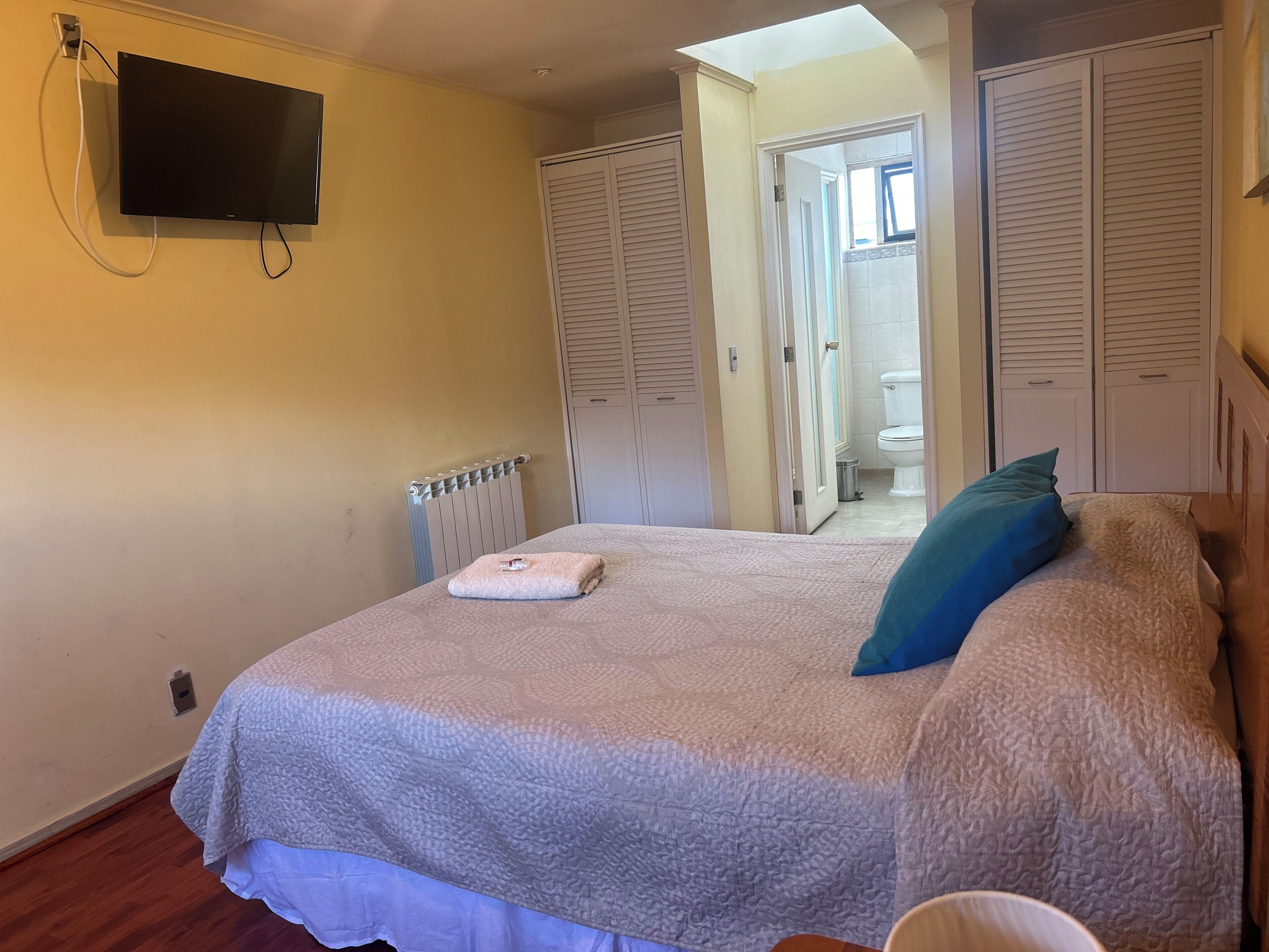 Quarto com cama, televisão fixada na parede, radiador e banheiro visível através de uma porta aberta.