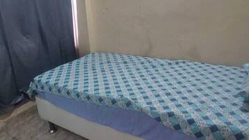 Um quarto pequeno com uma cama de solteiro, uma colcha estampada, cortina e chão de azulejos.