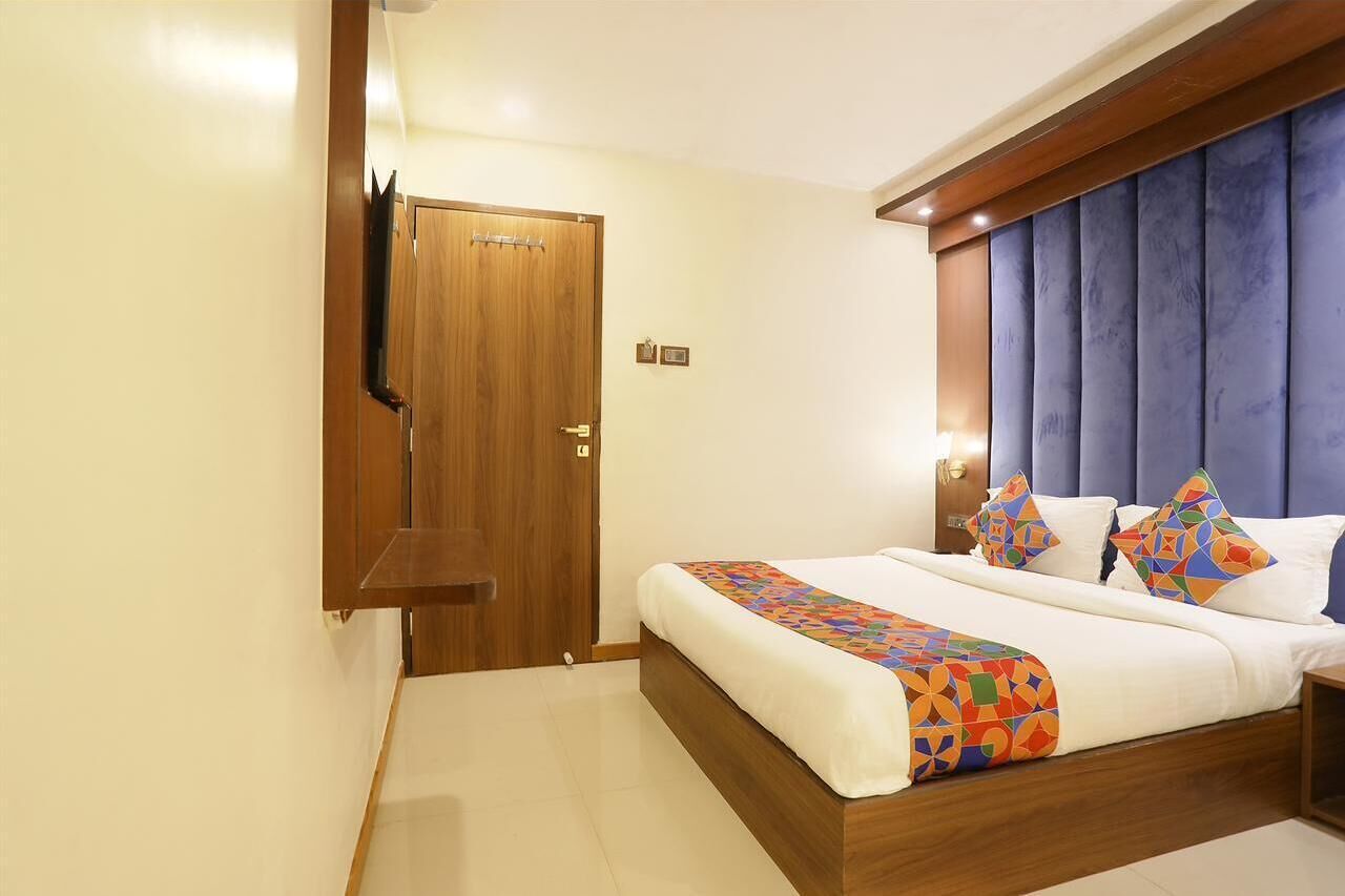 Quarto de hotel com porta de madeira, cama com colcha estampada em padrão geométrico colorido e cabeceira azul estofada.