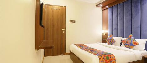Quarto de hotel com porta de madeira, cama com colcha estampada em padrão geométrico colorido e cabeceira azul estofada.