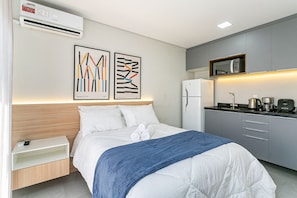 Quarto de hotel moderno com uma cama, uma mesinha de cabeceira, uma escrivaninha, uma cozinha compacta e duas obras de arte abstratas nas paredes.