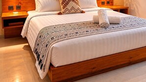 Quarto de hotel moderno com cabeceira de madeira, cama grande com roupa de cama estampada, duas mesas de cabeceira e arte nas paredes com folhagens de palmeira.