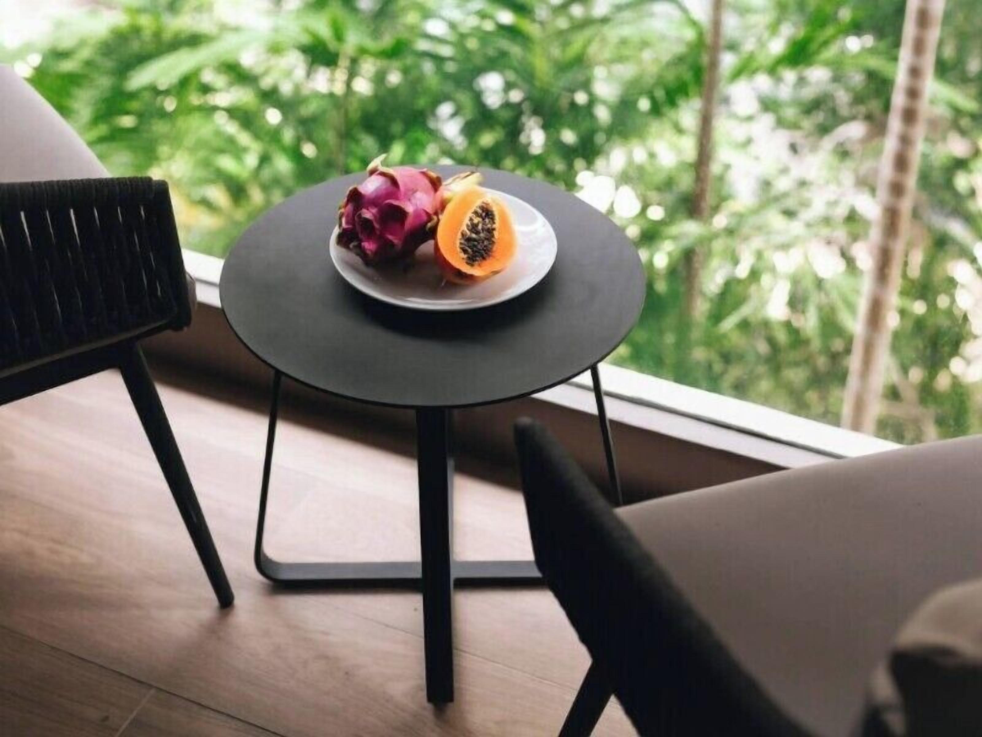 Uma mesa redonda com uma fruteira contendo pitaya e mamão, posicionada numa varanda com vista para o verde.