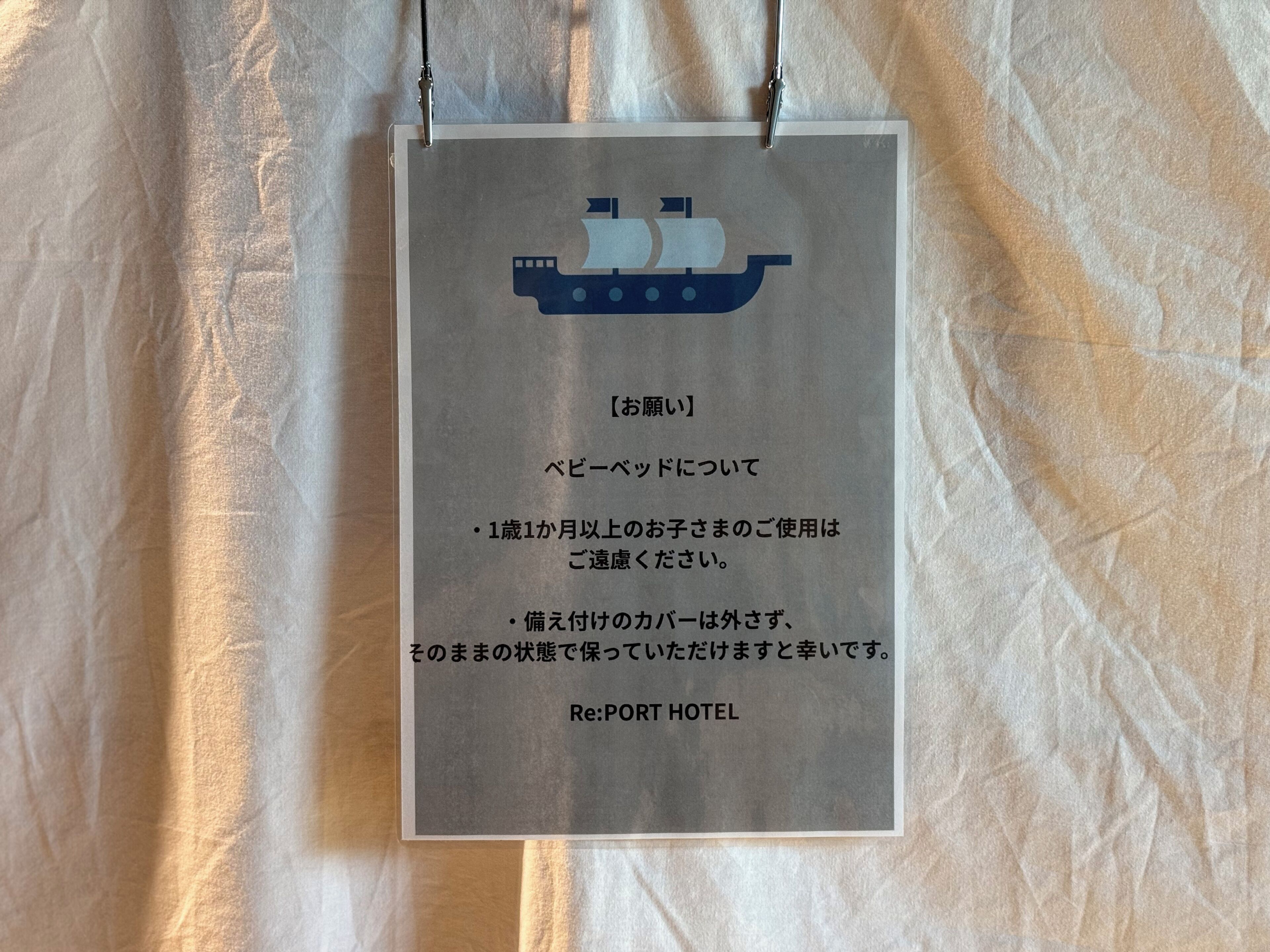 Um letreiro com ilustração de um navio e texto em japonês, pendurado em um fundo de tecido texturizado.
