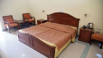 Quarto bem arrumado com uma cama grande, uma cadeira, uma mesinha de cabeceira com chaleira e uma pequena mesa com telefone.