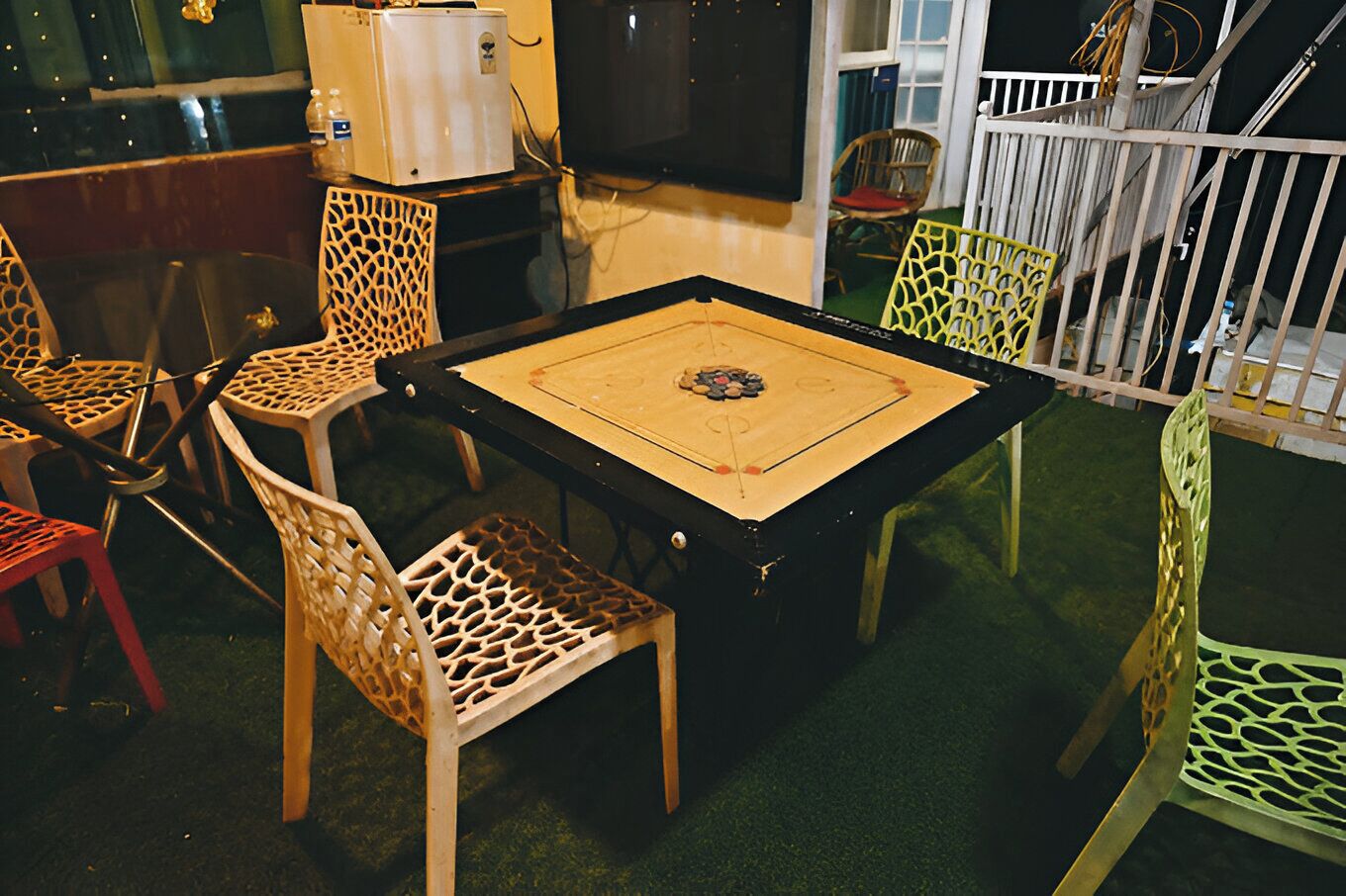 Uma mesa de bilhar com cadeiras ao redor, uma televisão e uma cerca branca ao fundo.