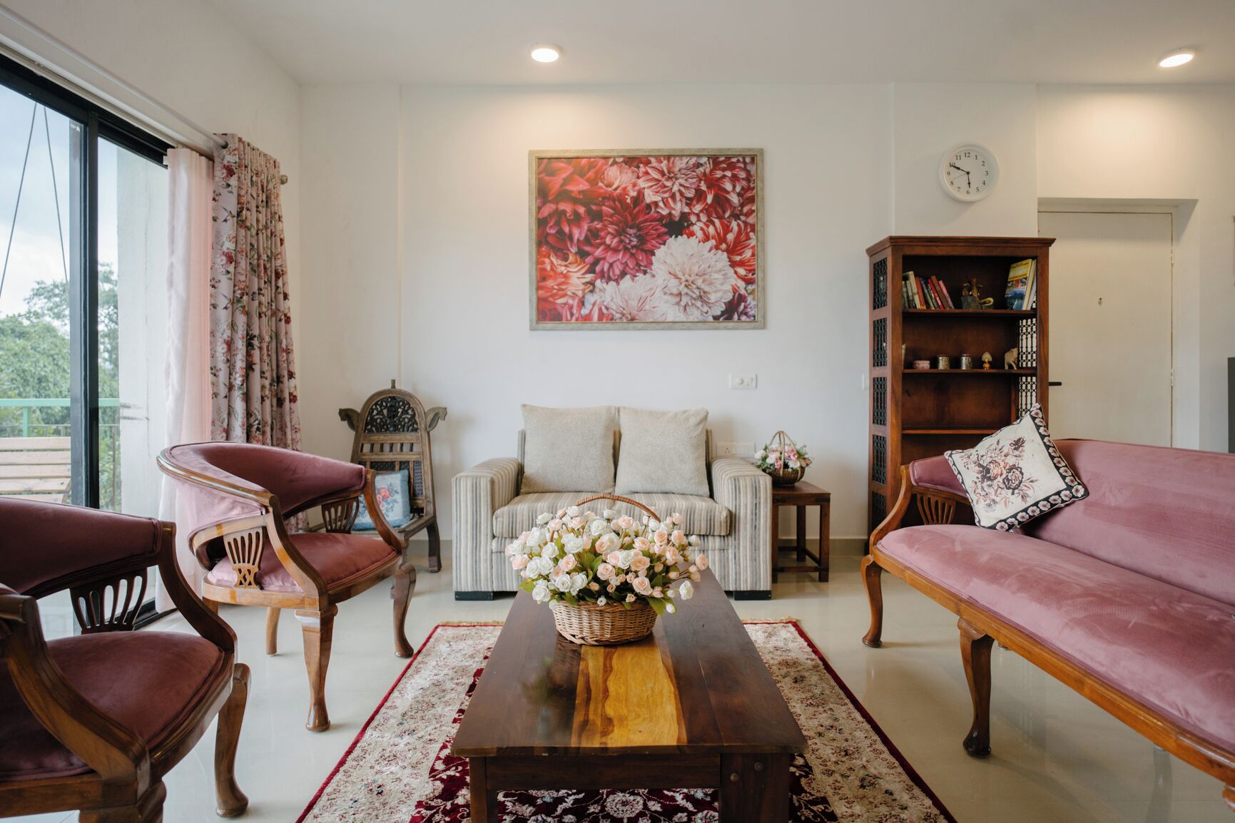 Uma sala de estar com sofá, poltronas, mesa de centro com arranjo floral, estante de livros e uma pintura floral grande na parede.