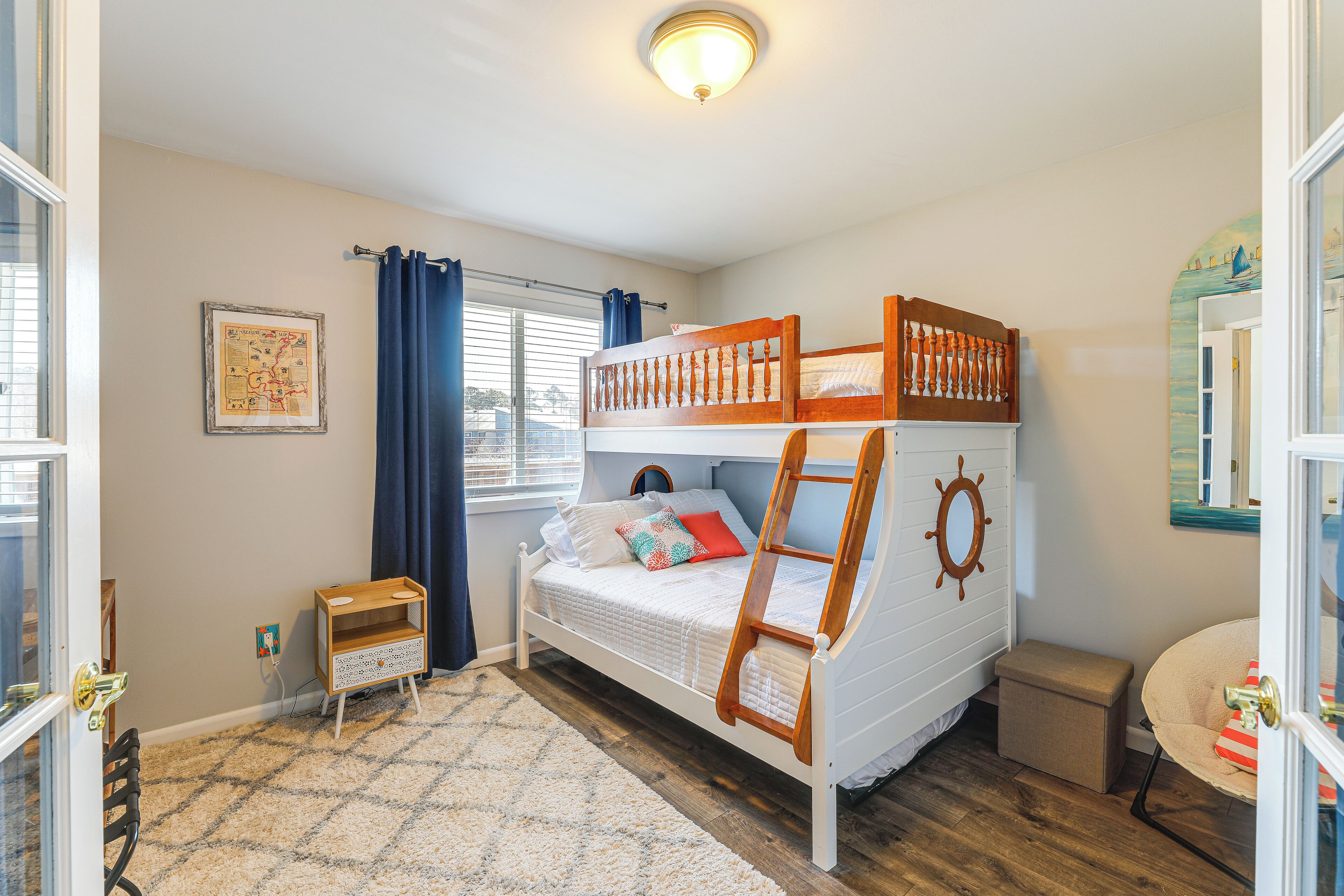 Uma cama de solteiro com decoração de roda de navio, um pequeno baú de madeira e uma cadeira com almofada listrada.