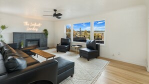 Uma sala de estar moderna com lareira, sofá de couro preto e vista para uma área residencial através de grandes janelas.