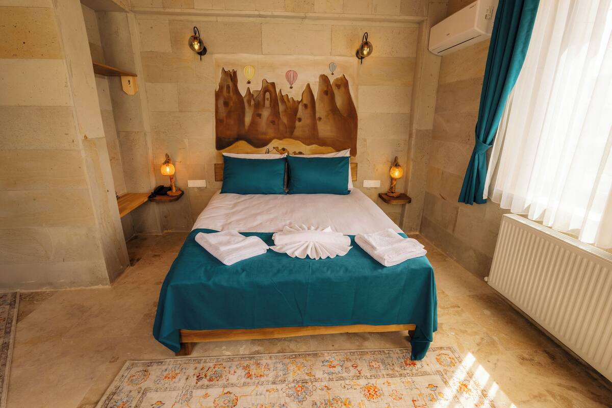 Cama com colcha verde-água, almofadas brancas e cabeceira decorativa. Há duas luminárias de mesa e uma janela com cortinas.