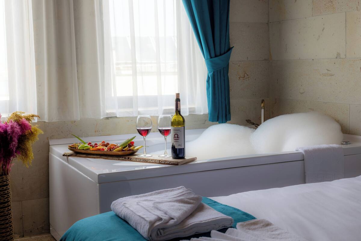 Quarto de hotel com cama, janela com cortinas, mesa com garrafa de vinho, copos e prato de fruta.