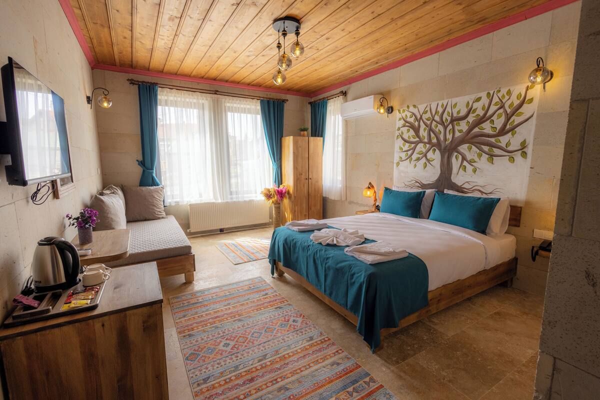 Quarto com teto de madeira, cama com roupa de cama azul, mesa de cabeceira com chaleira e televisão na parede.