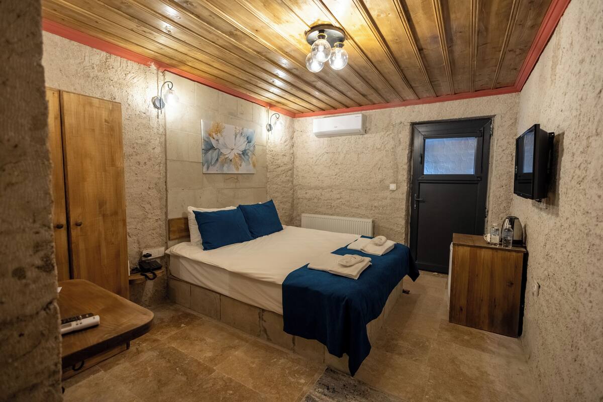Quarto com cama, mesa de cabeceira de madeira, uma pequena mesa, televisão e porta.