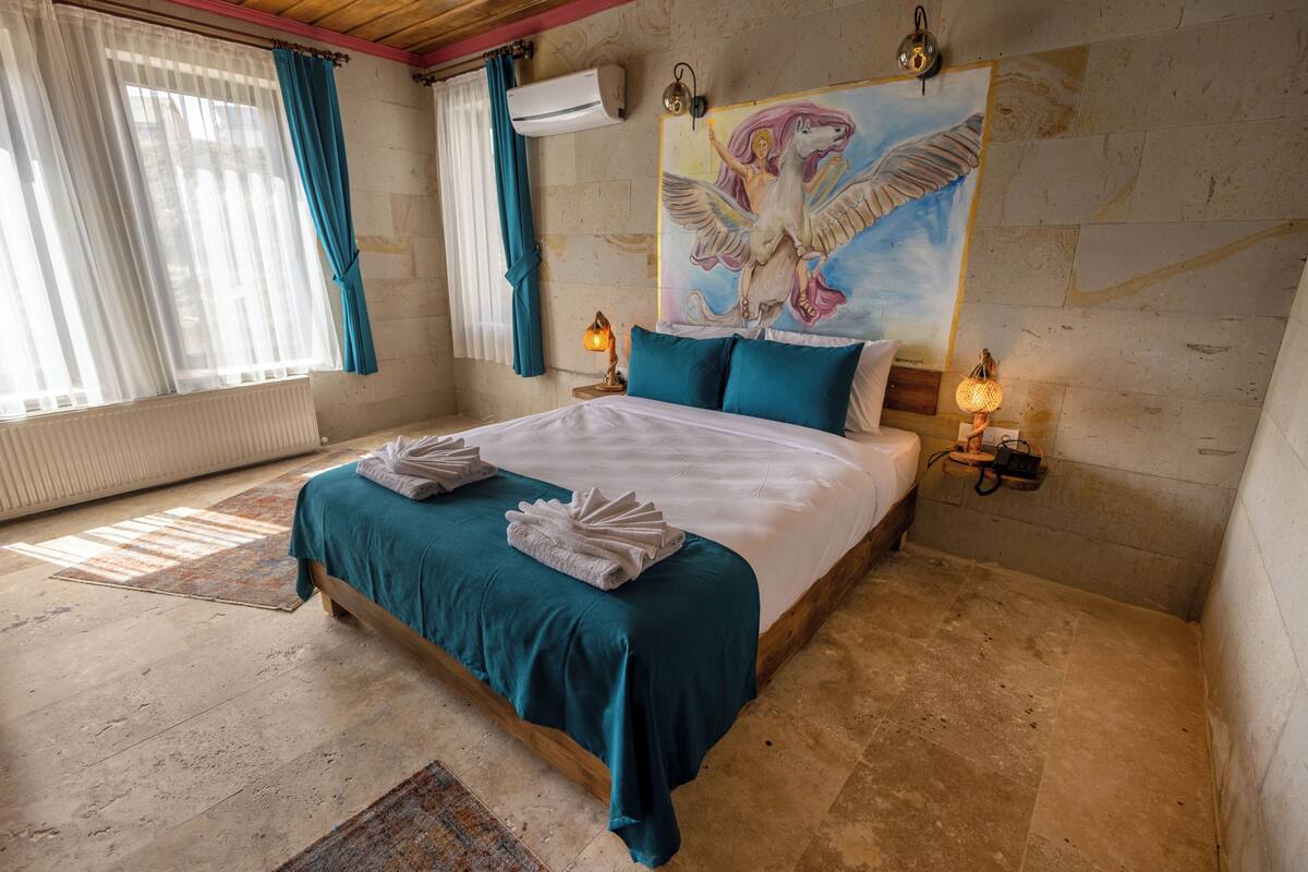 Quarto com uma cama grande, roupa de cama verde-água, um quadro de uma criatura mítica e uma parede de pedra.