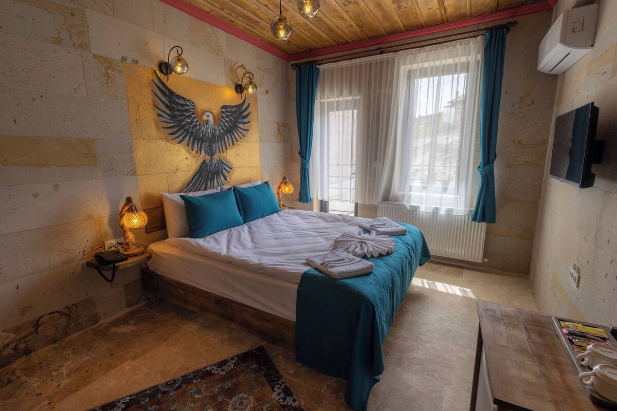 Quarto com cama grande, um objeto decorativo na parede, janela com cortinas e mesa de cabeceira com abajur.