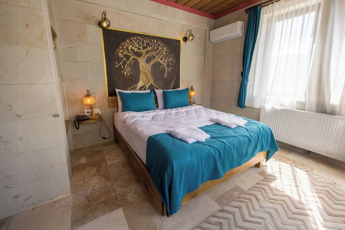 Quarto com cama, um quadro de árvore na parede, mesa de cabeceira com abajur e janela com cortinas.
