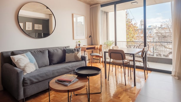 Uma sala de estar moderna com um sofá cinzento, uma mesa de centro redonda e um espaço de refeições com mesa e cadeiras. Um espelho grande e quadros emoldurados decoram as paredes.