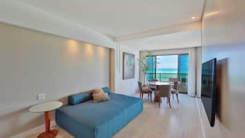 Uma sala de estar moderna com um sofá azul, uma mesa redonda e cadeiras. Há uma TV instalada na parede e uma janela com vista para o mar.