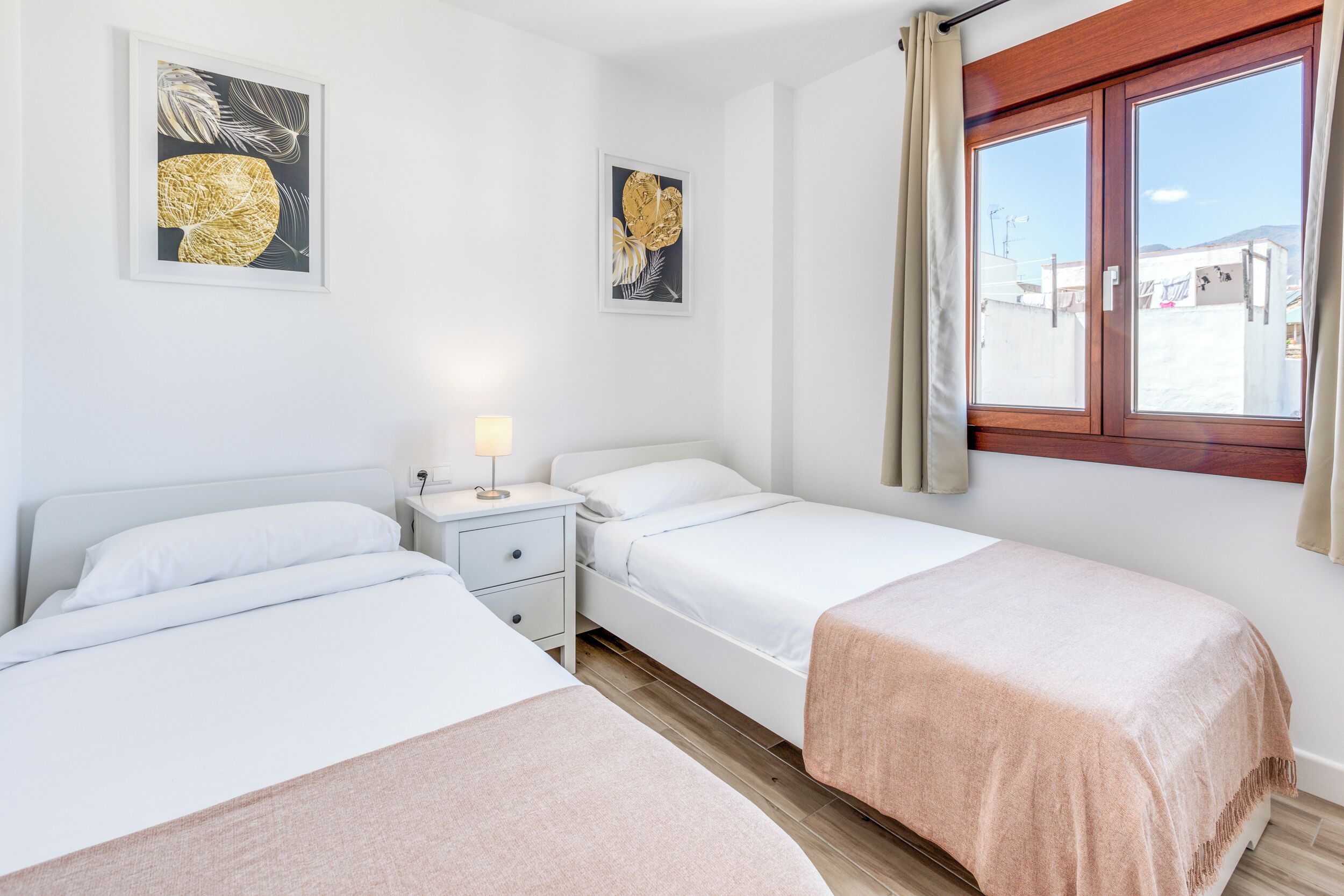 Quarto de hotel com duas camas de solteiro, uma mesa de cabeceira com abajur e uma janela com cortinas.