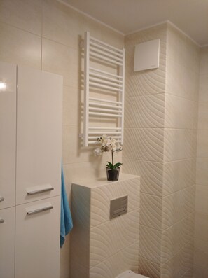 Uma casa de banho com armário branco, um porta-toalhas, uma pequena planta em vaso e um aquecedor fixado na parede.