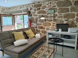 Uma sala de estar acolhedora com uma parede de pedra, um sofá com almofadas, uma mesinha de centro com abajur e uma estante com diversos objetos.