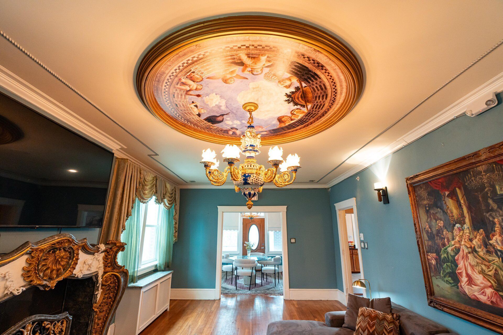 Um quarto com um lustre grande e ornamentado, uma sala de jantar com mesa redonda e cadeiras, e um quadro clássico grande na parede.