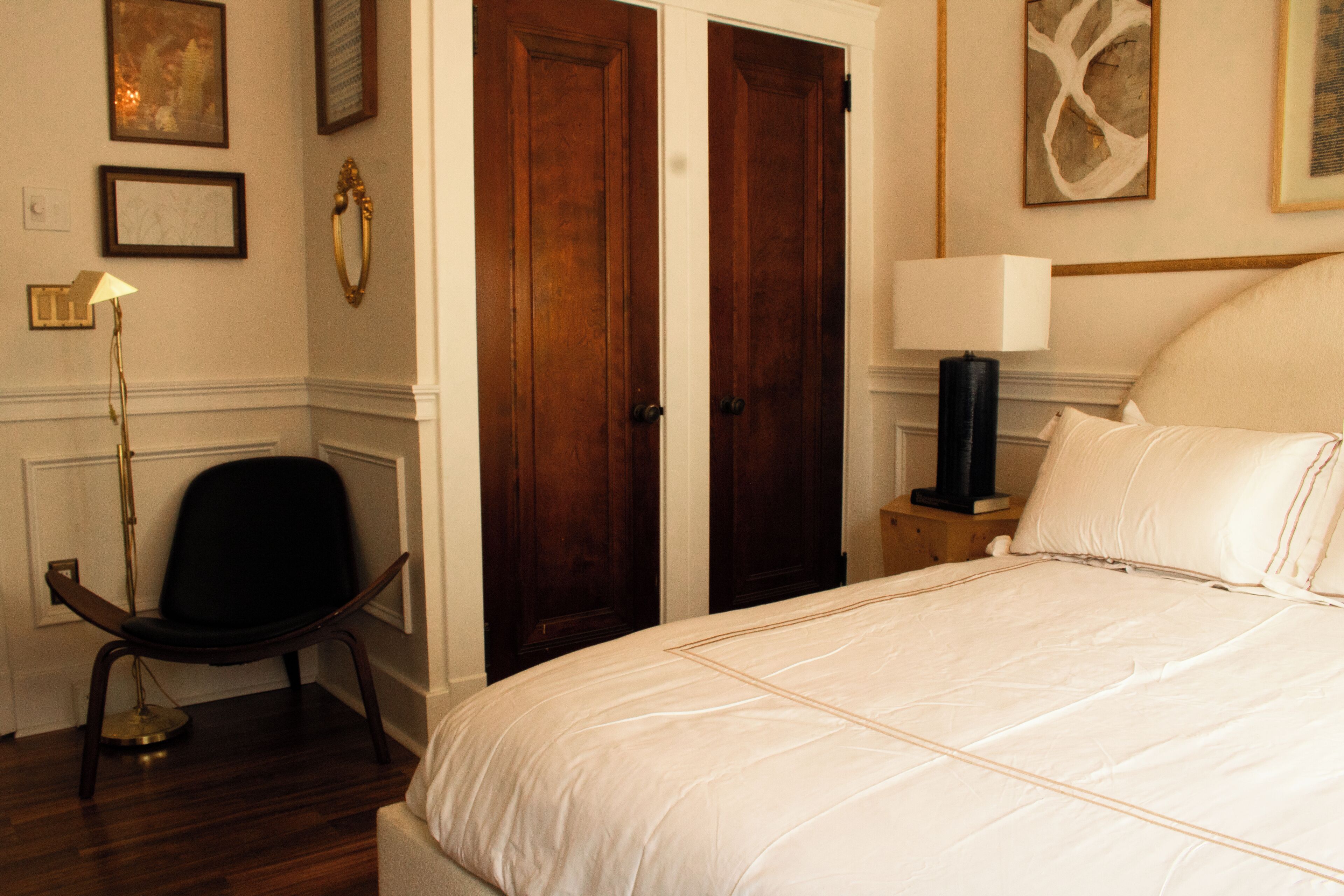 Quarto com uma cama grande, uma cadeira, escrivaninha com abajur e duas portas de madeira.