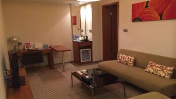 Quarto de hotel compacto com sofá, mesinha de centro, escrivaninha com cadeira, luminária e um quadro na parede.
