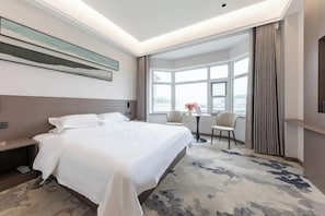 Quarto de hotel com uma cama grande, uma escrivaninha com cadeira, uma janela com vista e um quadro na parede.