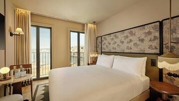 Quarto de hotel com uma cama grande, uma mesa de jantar, uma cadeira, uma mesinha de cabeceira com uma luminária e vista para a cidade pela janela.