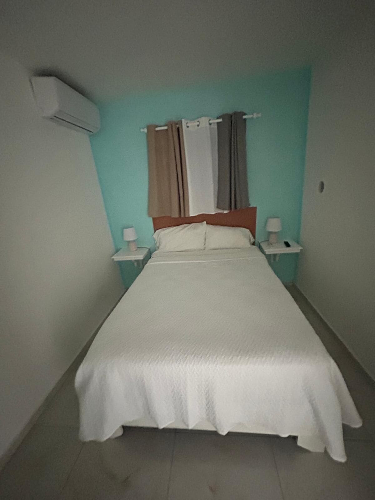 Quarto individual com uma colcha branca, duas mesas de cabeceira com abajures, um ar condicionado fixado na parede e uma parede na cor verde-água.