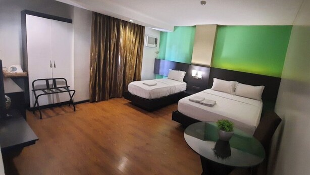Quarto de hotel com duas camas, uma escrivaninha, uma cadeira, uma mesinha com uma planta e uma cortina.