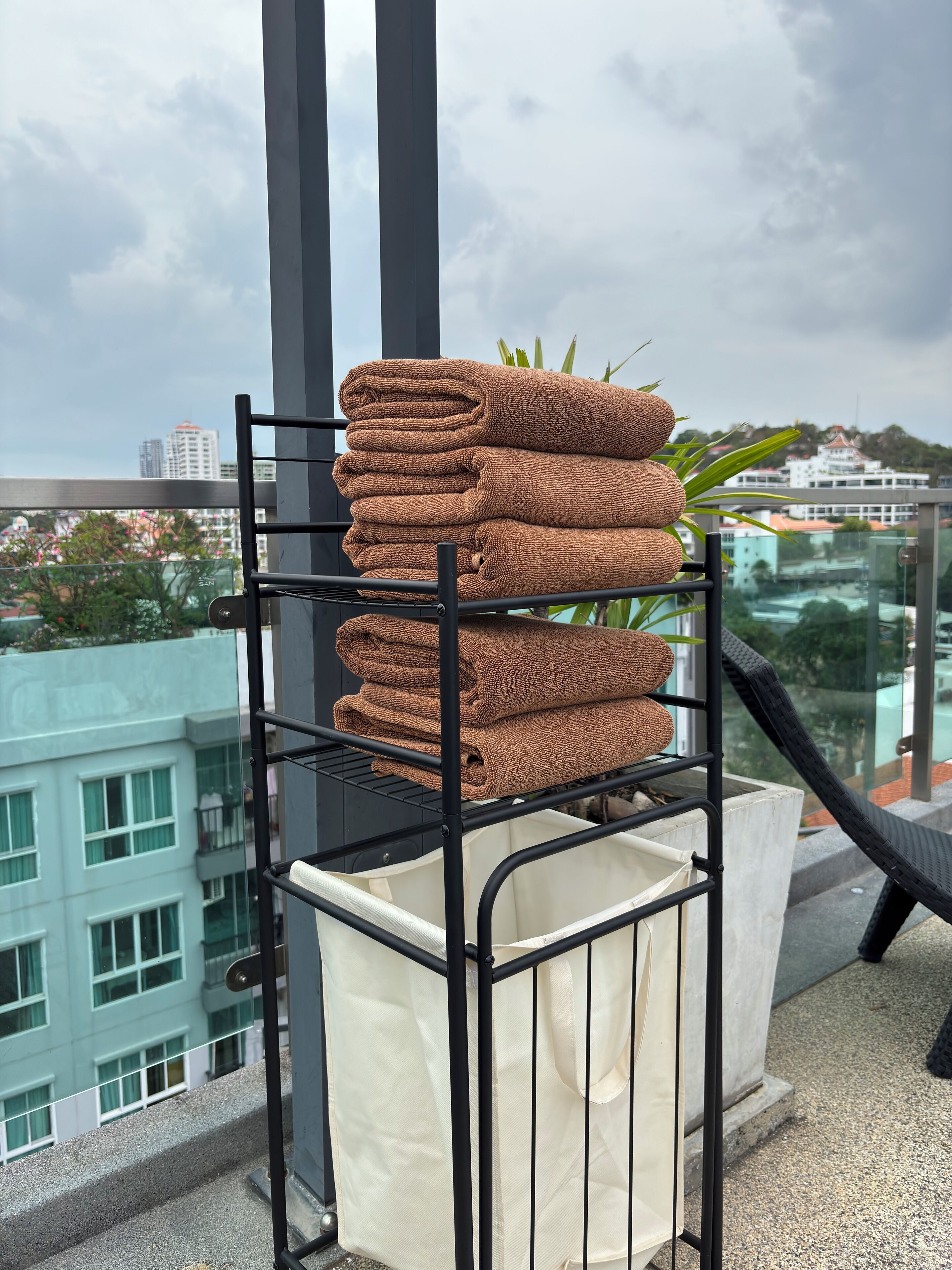 Uma estante de metal com toalhas castanhas empilhadas, uma lixeira branca e vista de prédios e árvores ao fundo.