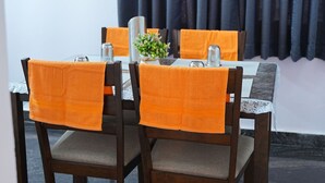 Mesa de jantar posta com quatro cadeiras, cada uma com uma toalha laranja. A mesa está coberta por um pano de mesa branco e há dois vasos sobre ela.
