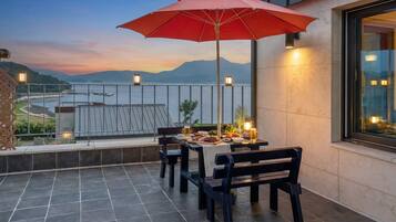 Terraço com mesa posta para refeições, um guarda-sol vermelho e vista para um corpo de água e montanhas.