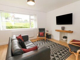 Uma sala de estar moderna com um sofá de couro preto, uma cadeira de madeira, um tapete estampado e uma TV fixada na parede.