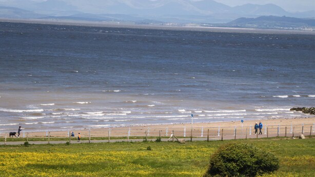 Vista costeira com praia arenosa, primeiro plano com grama e montanhas ao longe.