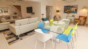 Uma sala de estar moderna com uma mesa de jantar de vidro, cadeiras brancas e azuis, um sofá e uma televisão.