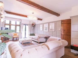 Uma sala de estar ampla com um sofá grande, uma poltrona menor e uma mesa de centro. Há uma porta de madeira à direita e uma janela com cortinas à esquerda.