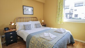 Uma cama bem arrumada com almofadas estampadas, uma luminária de cabeceira, um quadro na parede e uma janela com cortinas.